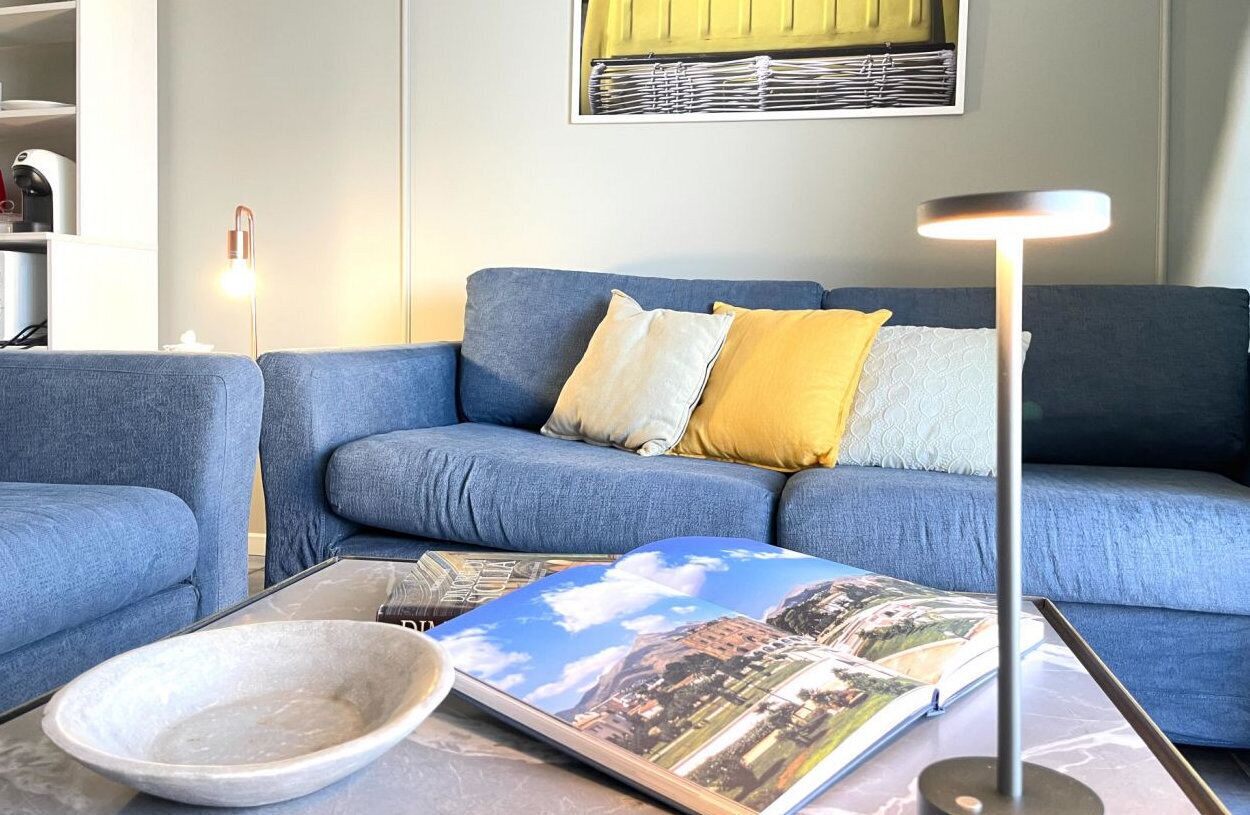 Uma sala de estar com um sofá azul, uma mesa de centro com um livro e um abajur.