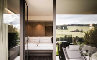 Quarto moderno com uma cama grande, painéis de madeira e uma varanda com mobiliário de exterior.