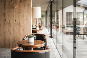 Uma área de estar moderna ao ar livre, com uma mesa e cadeiras de madeira, uma planta em vaso e um muro de vidro que oferece vista do interior.