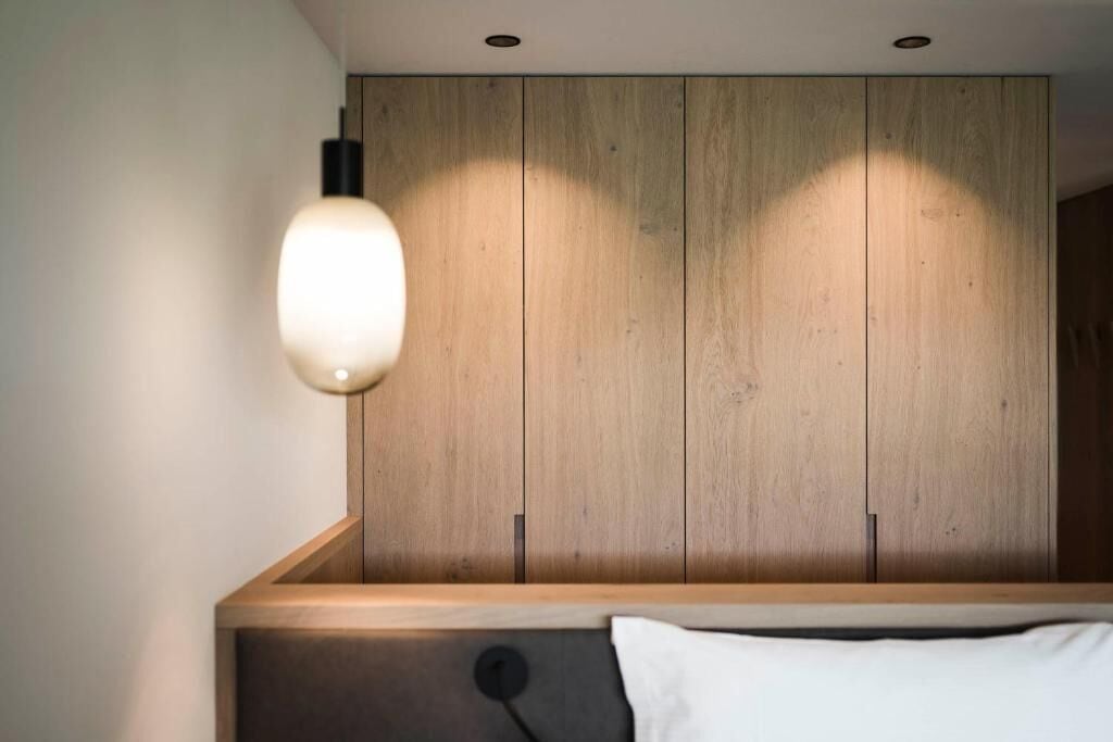 Quarto com cabeceira de madeira, travesseiro branco e luminária fixada na parede.