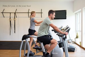 Duas pessoas se exercitando em bicicletas ergométricas numa academia, com uma frase motivacional na parede.