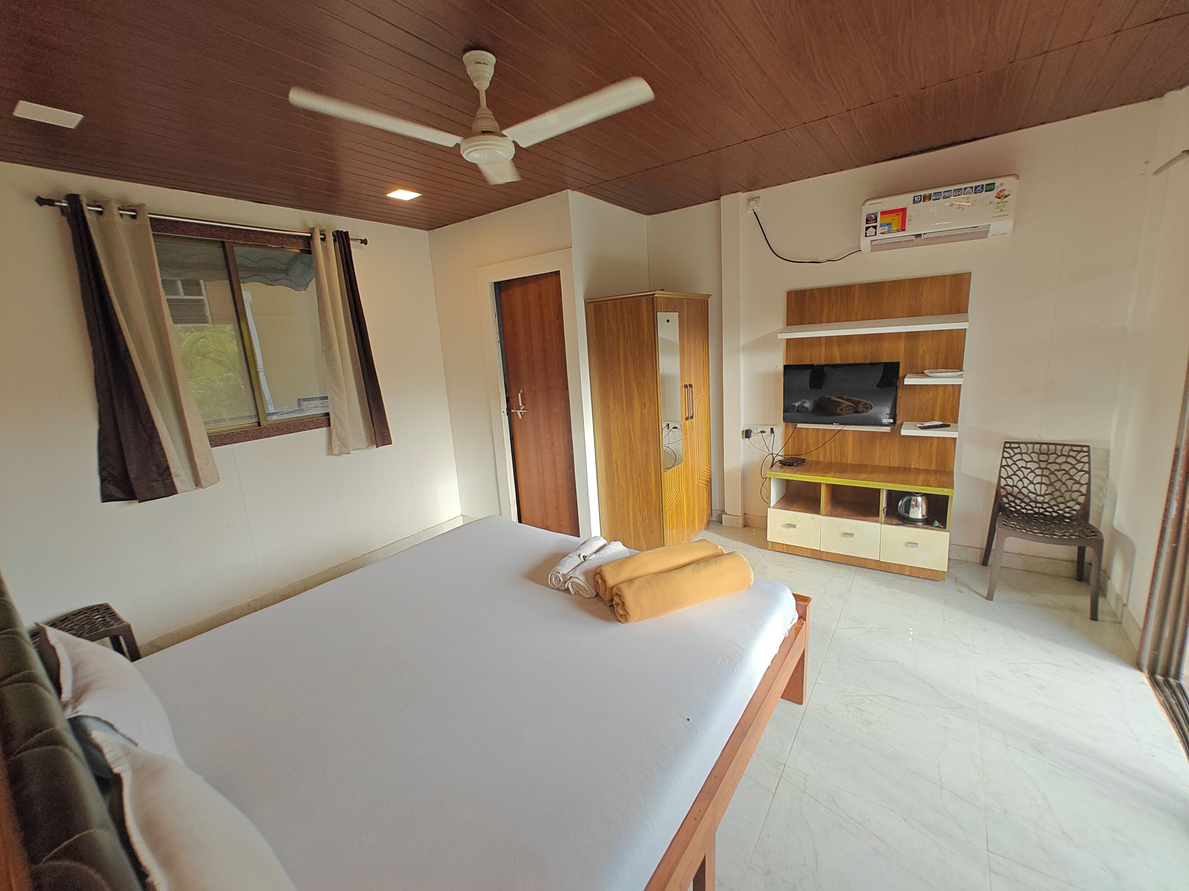 Quarto de hotel com cama, ventilador de teto, armário, suporte para televisão e uma cadeira.