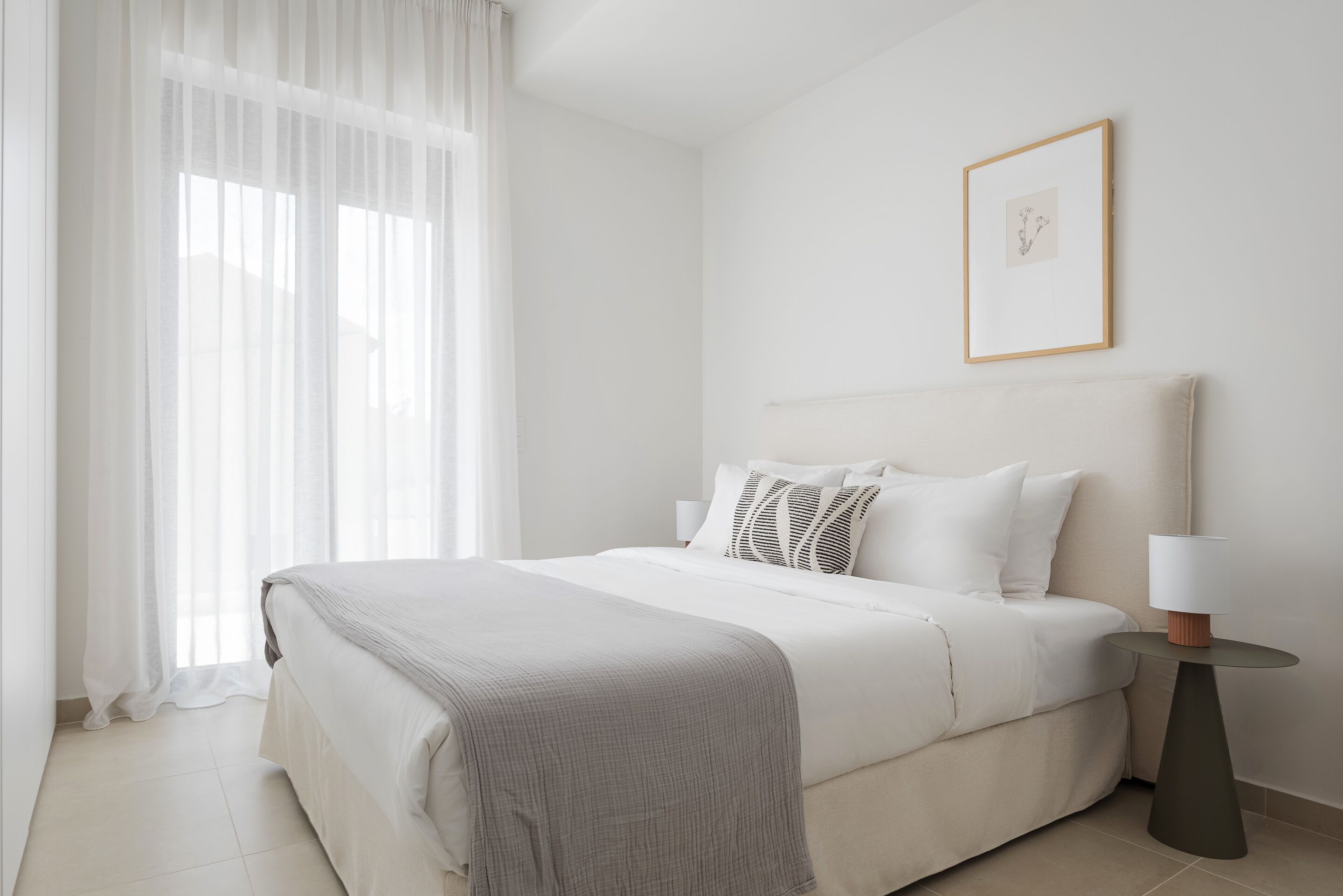 Quarto com cama, mesa de cabeceira com abajur, janela com cortinas leves e um quadro na parede.