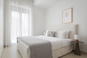 Quarto com cama, mesa de cabeceira com abajur, janela com cortinas leves e um quadro na parede.
