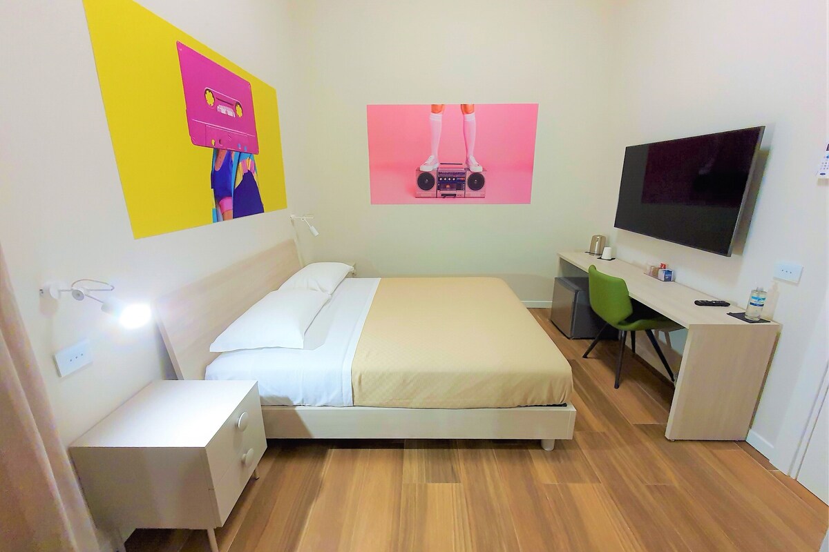 Quarto de hotel com cama, mesa de cabeceira, escrivaninha, cadeira e televisão.