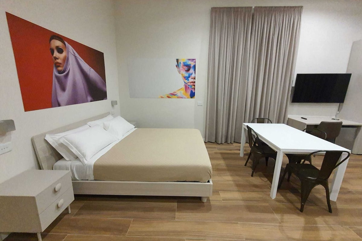 Quarto de hotel com cama, escrivaninha, cadeira, televisão e duas pinturas na parede.