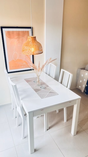 Uma mesa de jantar branca com uma luminária trançada, cadeiras brancas e uma obra de arte emoldurada na parede.