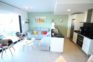 Uma sala de estar moderna com sofá, cadeiras e mesa de jantar. Há uma área de cozinha com eletrodomésticos e geladeira.