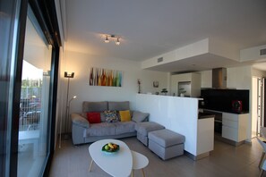 Uma sala de estar moderna com um sofá cinzento, uma mesa de centro branca e uma cozinha ao fundo.