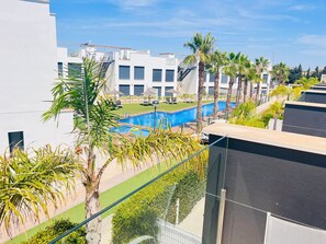 Um edifício moderno com piscina no terraço, rodeado por palmeiras e um céu azul límpido.