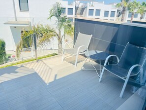 Uma varanda com área de estar, uma pequena palmeira e um edifício branco ao fundo.