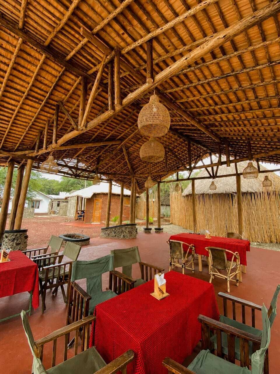 Uma área de refeições ao ar livre com mesas e cadeiras, um telhado de palha e vista para algumas cabanas tradicionais ao fundo.