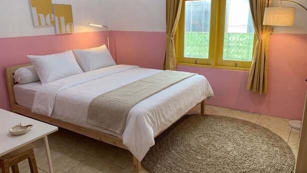 Quarto com cama, mesa de cabeceira, luminária, janela com cortinas e vista para o verde lá fora.