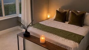 Quarto com cama, almofadas, mesa de cabeceira com uma vela e janela com cortinas.