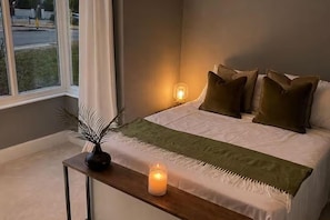 Quarto com cama, almofadas, mesa de cabeceira com uma vela e janela com cortinas.