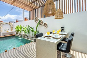 Uma área de refeições à beira da piscina, com uma mesa posta para dois, rodeada por vasos de plantas e vista para a piscina.