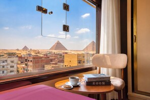 Quarto de hotel com vista para pirâmides, uma pequena mesa com uma xícara de café, um livro intitulado 'This is Home' e uma cadeira.