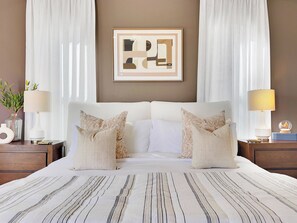 Quarto de hotel com uma cama grande, duas mesas de cabeceira, uma obra de arte abstrata emoldurada acima da cama e cortinas leves nas janelas.