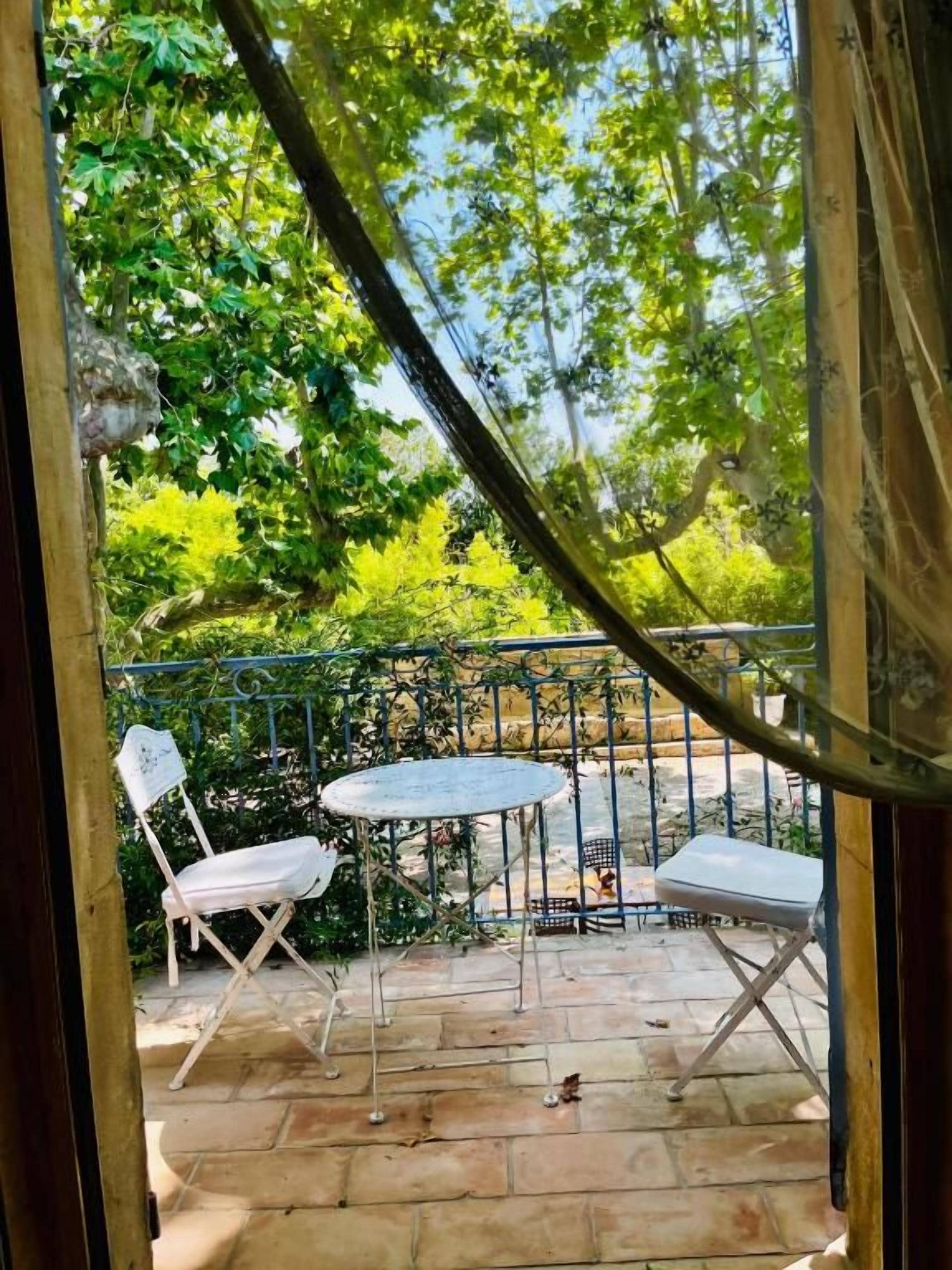 Pátio externo com mesa e duas cadeiras, cercado por um portão de metal e vegetação exuberante.