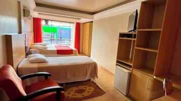 Quarto de hotel com cama, escrivaninha, cadeira, televisão e guarda-roupa.