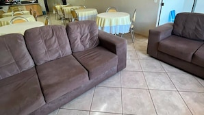 A spacious room with a large brown sofa, a dining area with round tables, and a kitchen area in the background.