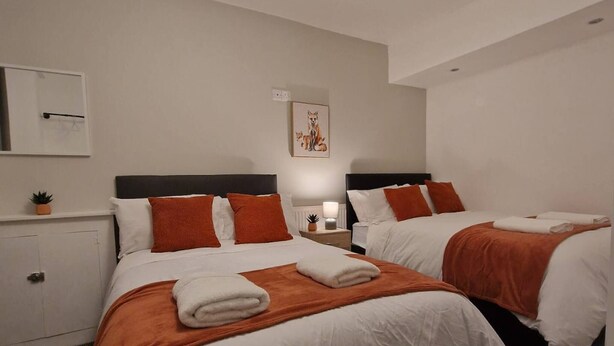 Quarto de hotel com duas camas, roupa de cama branca, almofadas de laranja e luminária branca, um quadro na parede e um lustre.