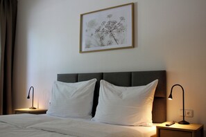 Quarto de hotel com cama, duas luminárias de cabeceira, mesa de cabeceira, quadro na parede e tomada.