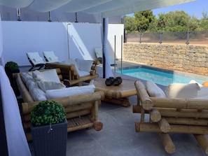 Área de piscina com espreguiçadeira de bambu, almofadas brancas e um muro de pedra ao fundo.