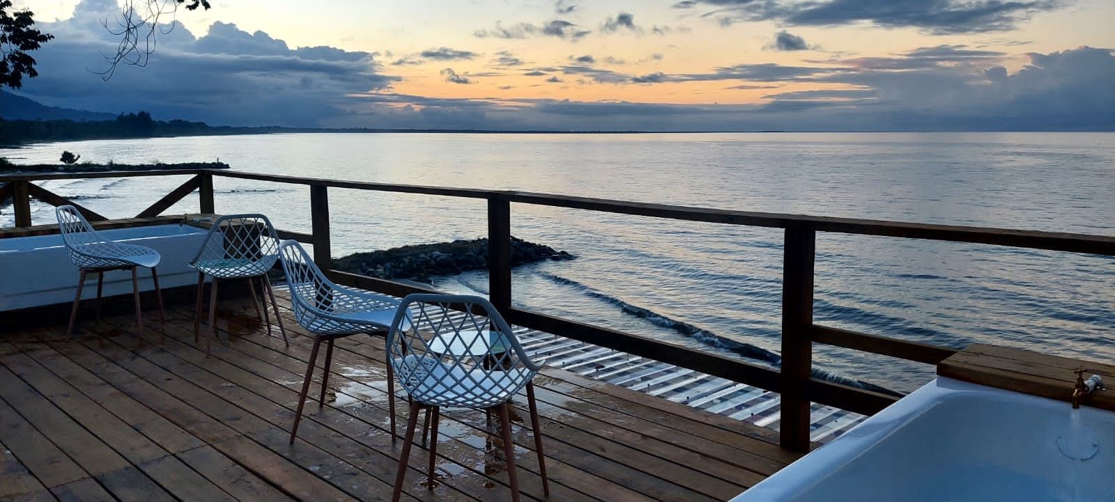 Um deck de madeira com duas cadeiras brancas e uma mesa, com vista para uma massa de água calma.