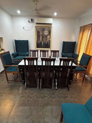 Uma área de refeições com uma mesa retangular rodeada por cadeiras de madeira, um ventilador de teto e um ar condicionado fixado na parede.
