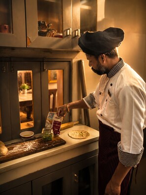 Um cozinheiro prepara alimentos em uma cozinha com eletrodomésticos de aço inoxidável e vista para uma sala de jantar.