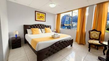 Quarto de hotel com uma cama grande, uma cadeira, uma mesinha e uma janela com cortinas.