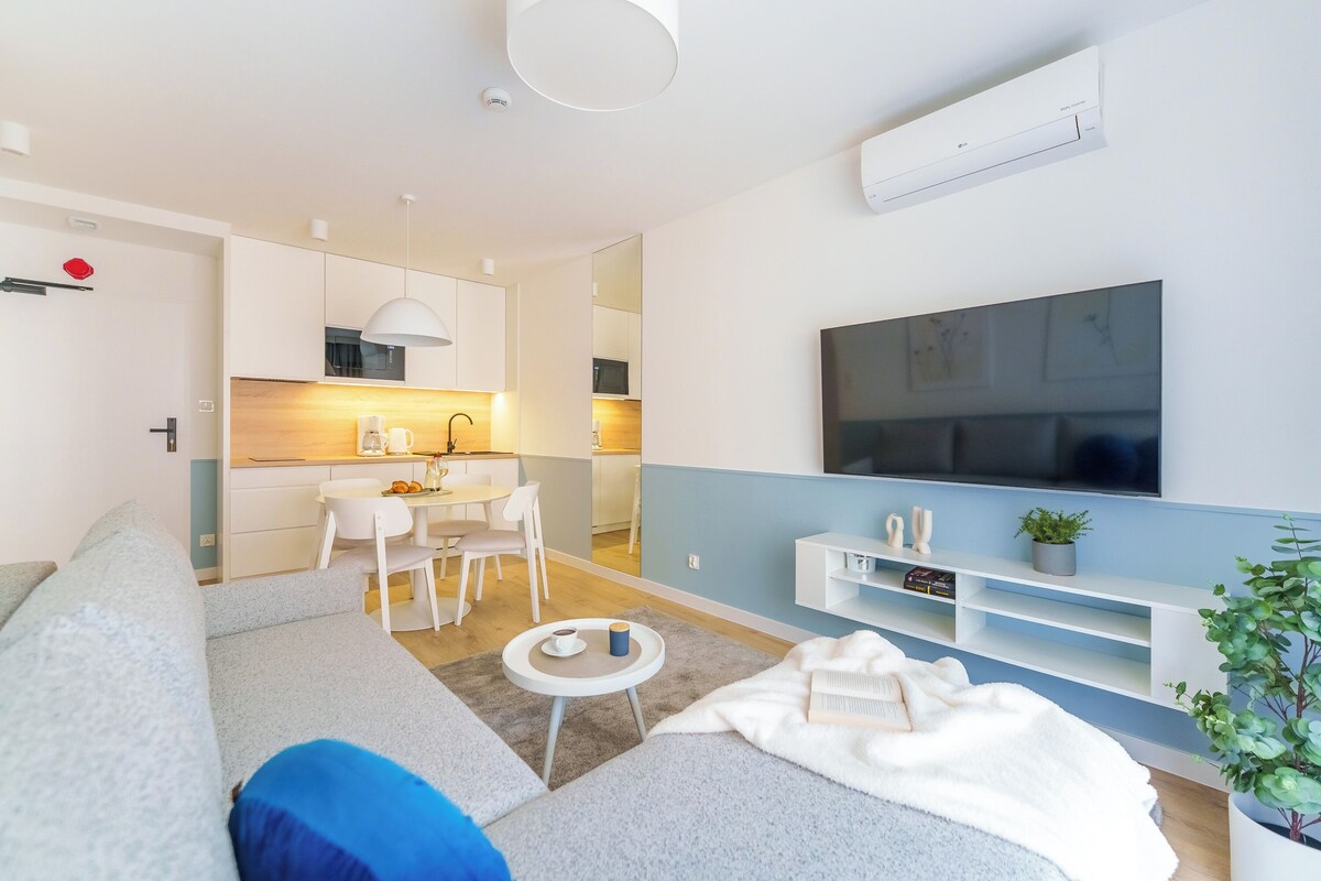 Uma sala de estar moderna com sofá, uma mesa de centro, uma TV e uma área de cozinha ao fundo.