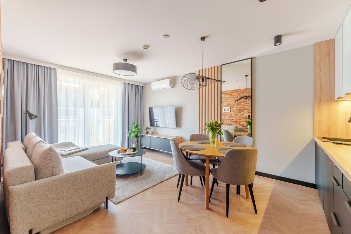 Uma sala de estar moderna com sofá, mesa de jantar e cadeiras, área de cozinha com bancada e uma grande janela com cortinas.
