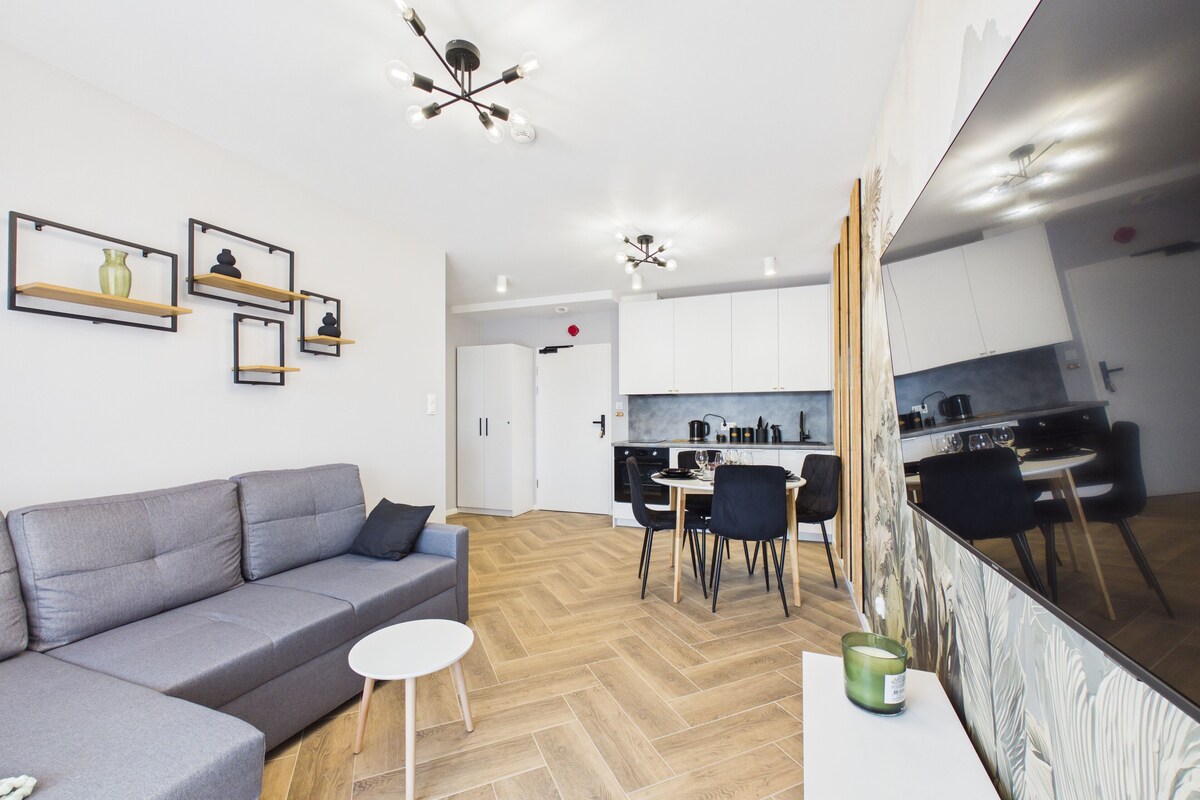 Uma sala de estar moderna com um sofá cinza, uma mesa pequena branca e um piso de madeira em padrão herringbone.