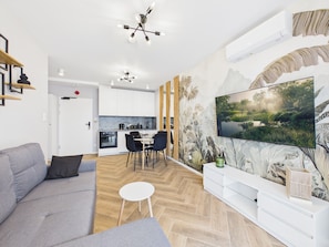 Uma sala de estar moderna com um sofá cinzento, uma área de jantar com mesa e cadeiras, uma cozinha com armários brancos e uma TV grande fixada na parede.