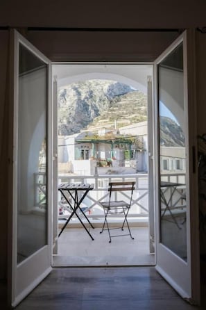 Uma porta aberta dá para uma varanda com mesa e cadeira, com vista para uma paisagem montanhosa.