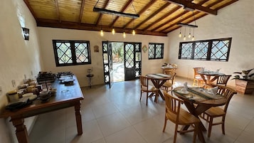 A spacious dining area with wooden tables and chairs, a kitchen counter with various items, and large windows with cross ventilation.