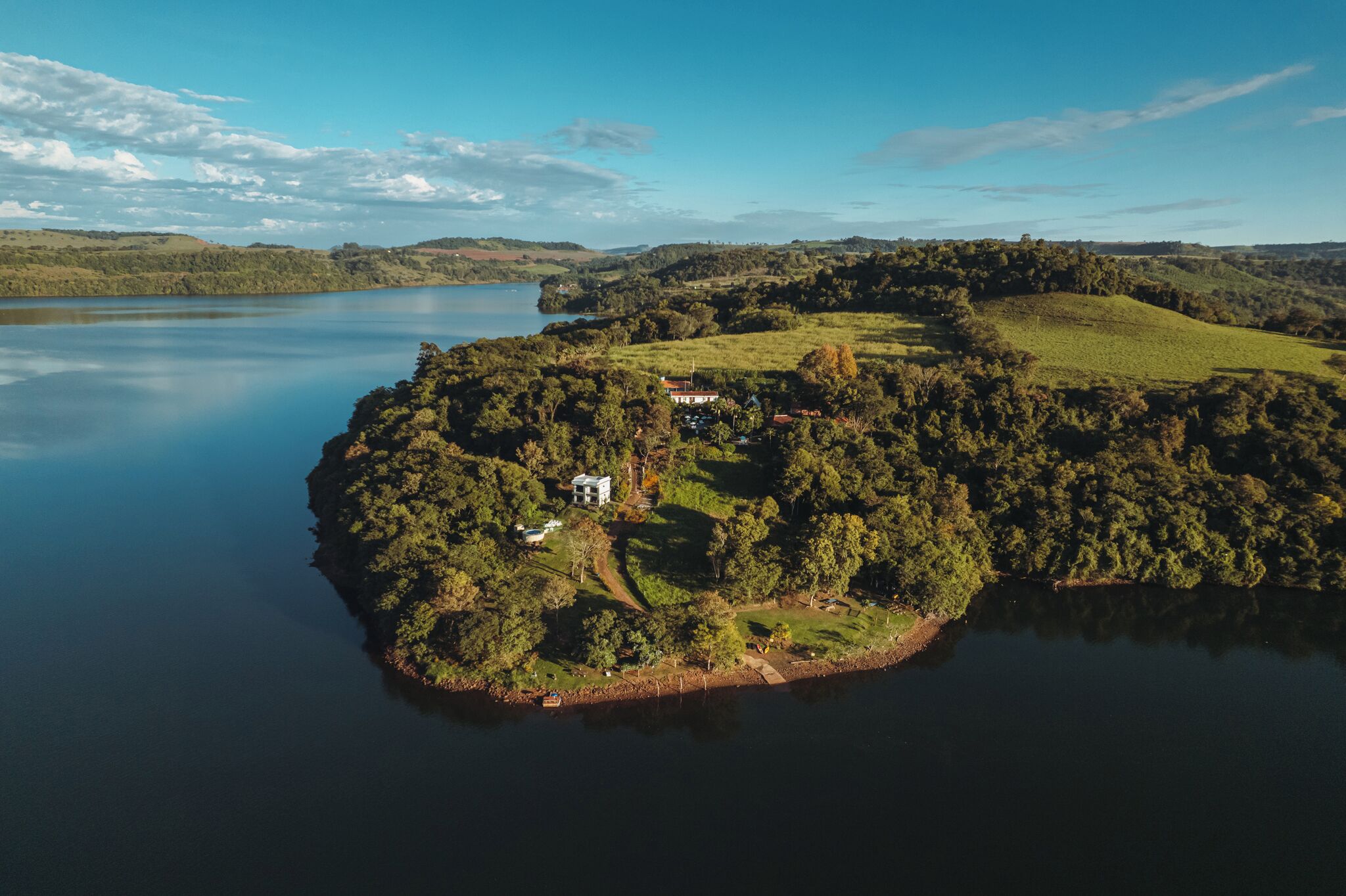 Vista aérea de uma propriedade à beira de um lago, com uma casa rodeada por uma floresta densa, céu claro e águas calmas.
