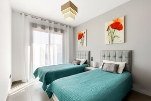 Quarto de hotel com duas camas, uma janela grande com cortinas translúcidas e obras de arte nas paredes.