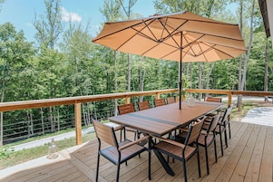 Um deck de madeira com mesa, cadeiras, um guarda-sol e vista para uma floresta.