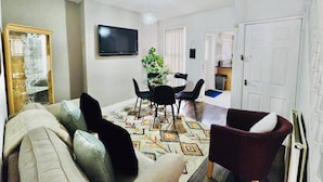 Uma sala de estar com sofá, uma mesa de jantar com cadeiras, uma televisão na parede e uma porta que dá para a cozinha.