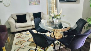 Uma sala de estar com uma mesa de vidro redonda, cadeiras pretas, um sofá bege, uma televisão e um quadro na parede.