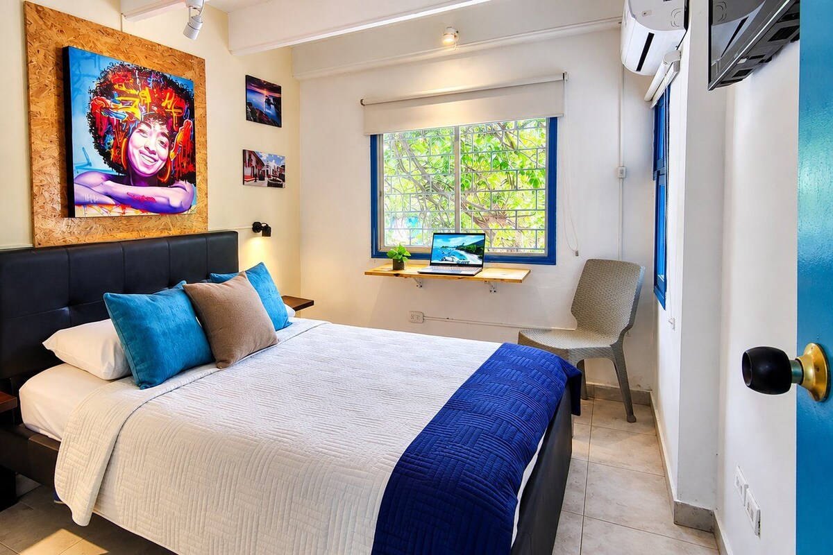 Quarto com cama, escrivaninha, laptop, cadeira e janela vista para árvores.