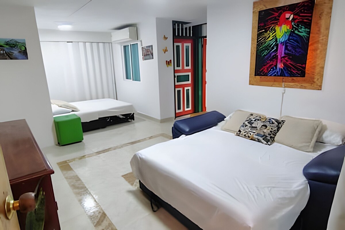 Quarto de hotel com cama, sofá, lixeira verde e um quadro colorido na parede.