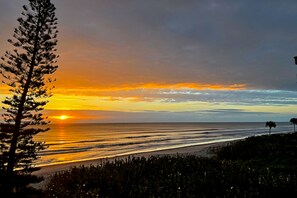 Pôr do sol sobre uma praia com ondas, uma árvore e uma cerca.