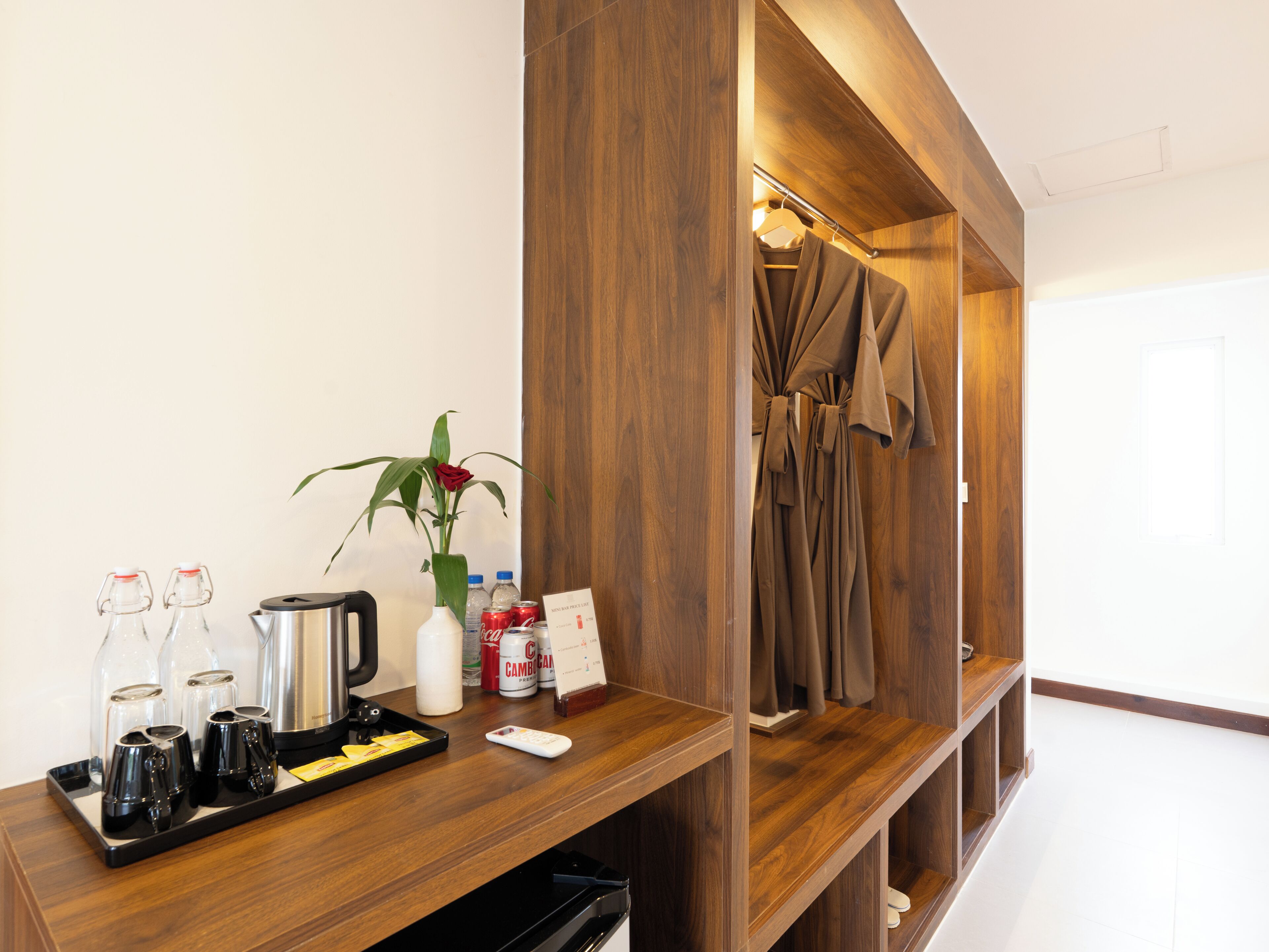 Quarto de hotel com toucador de madeira, roupão pendurado em um suporte, bandeja com xícaras de café, vaso com planta e uma garrafa de Coca-Cola.