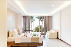 Quarto de hotel moderno com cama, sofá, poltrona e vista para árvores através de grandes janelas.
