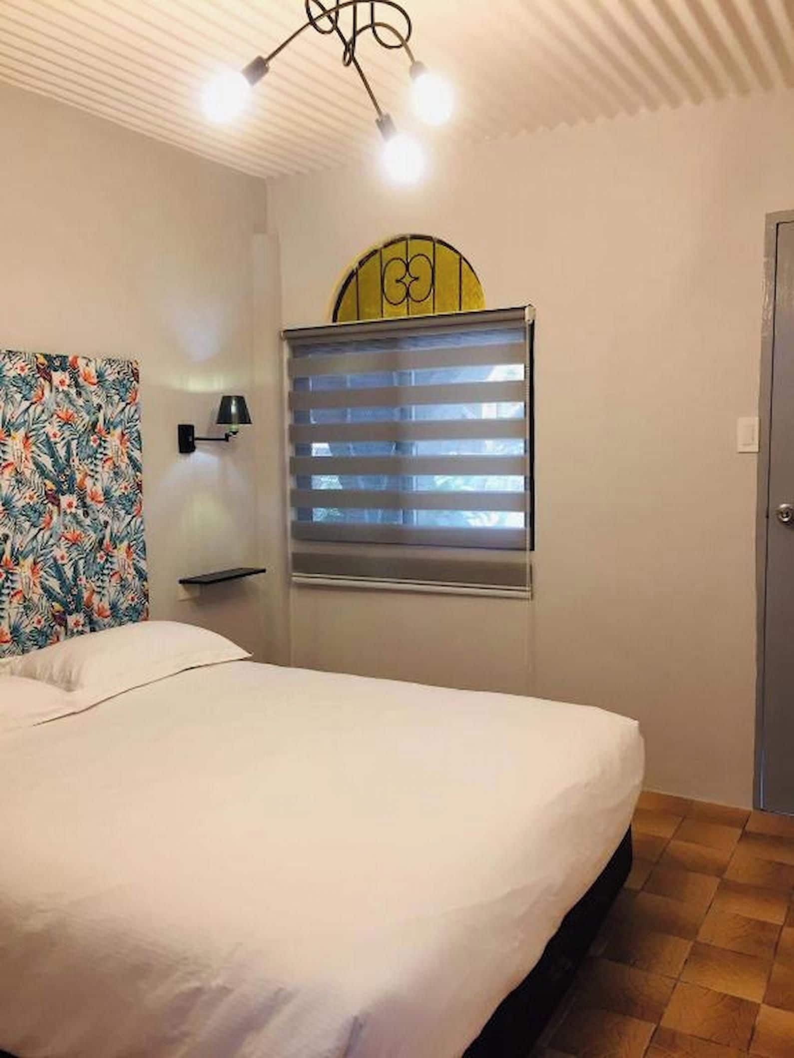 Quarto com cama, janela com persianas, luminária na parede e porta.