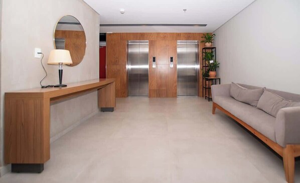 Um hall moderno com balcão de recepção, espelho redondo, sofá, dois elevadores e planta em vaso.